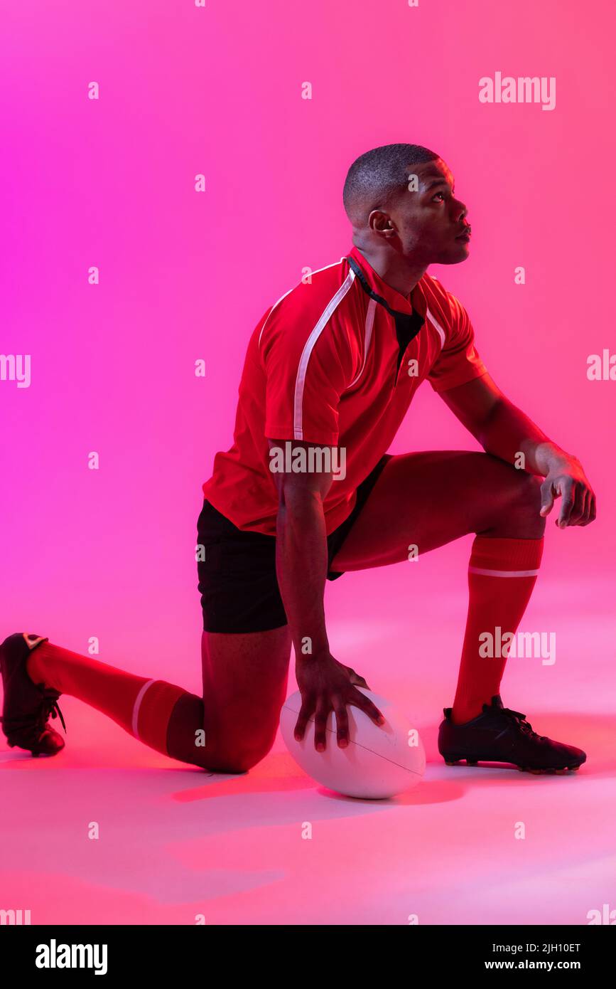 African american male rugby player crouching with rugby ball over pink ...