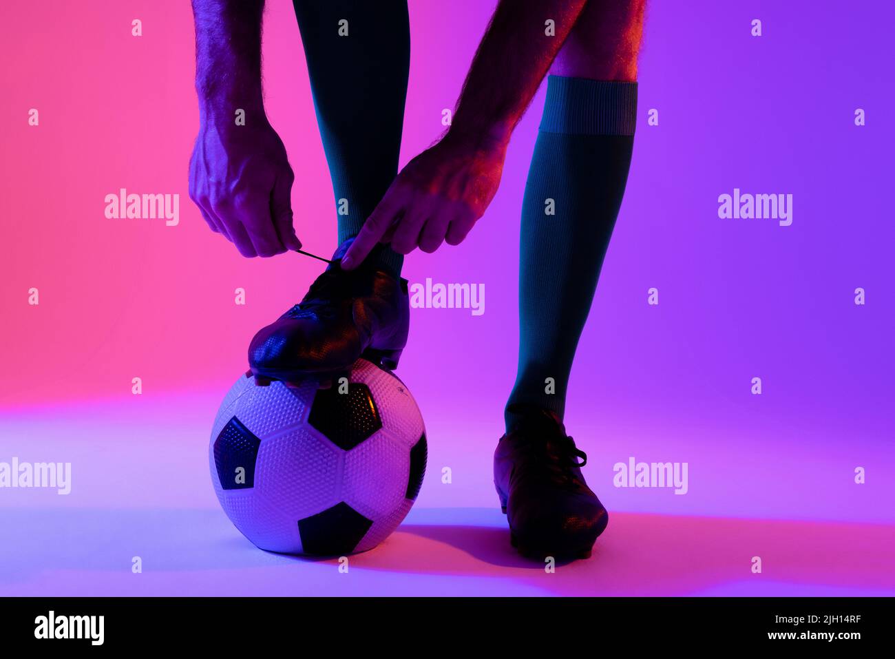 Caucasian male soccer player with football tying shoes over neon pink ...