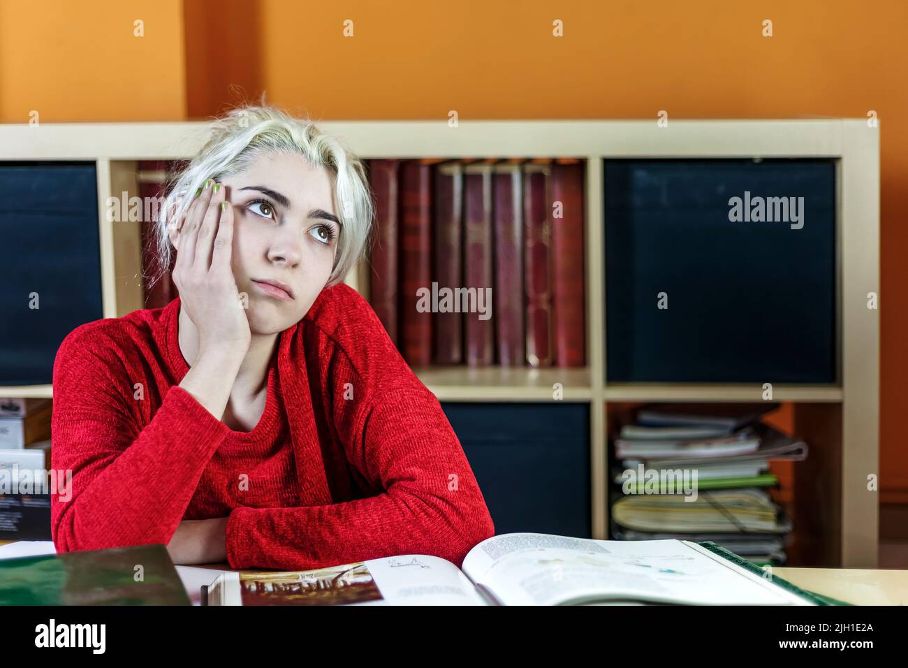 Bored female nerd student studies all day, surrounded with pile of books, keeps hand under chin ...