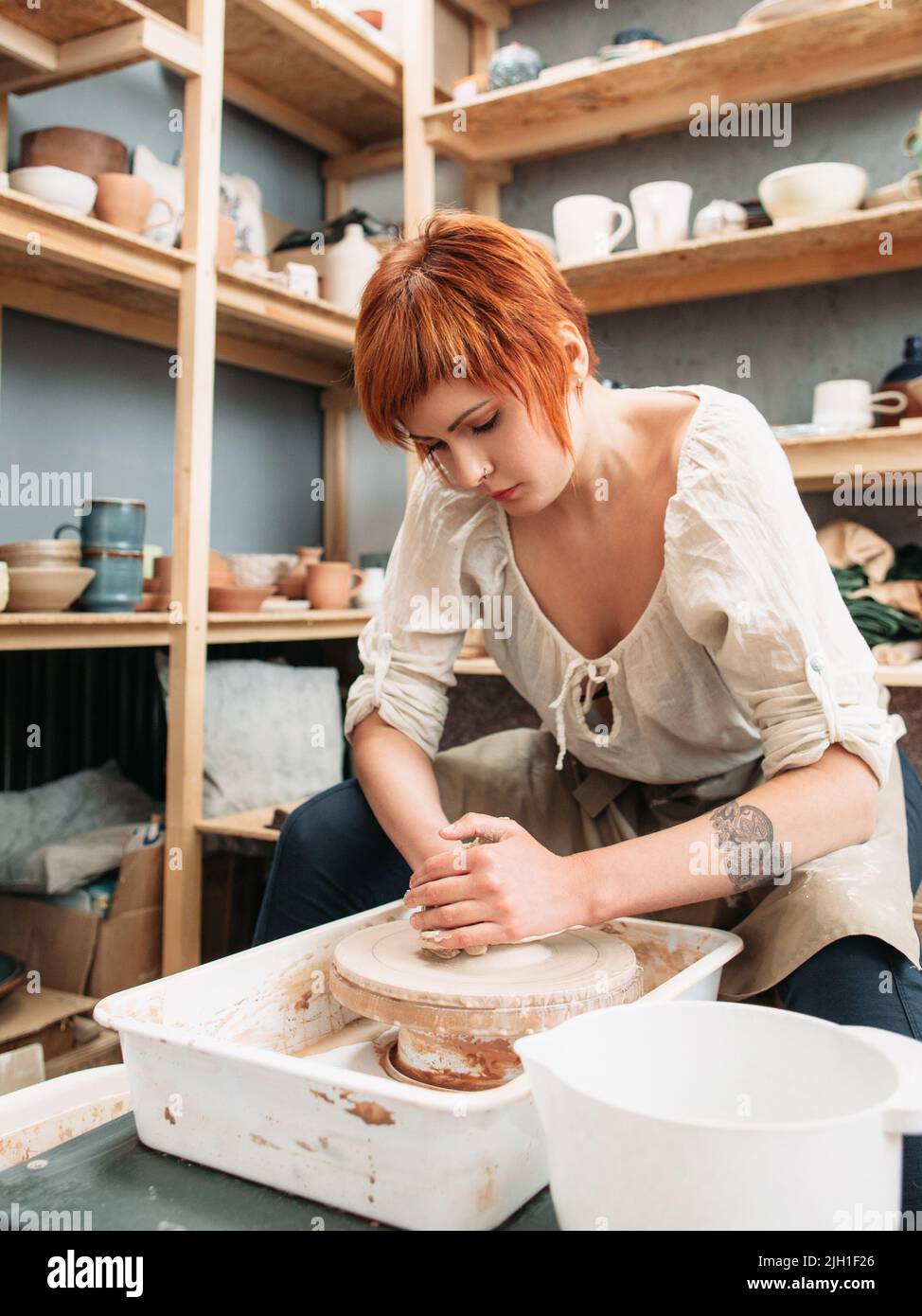 Woman works on potters wheel at studio. Plenty of pottery on background ...