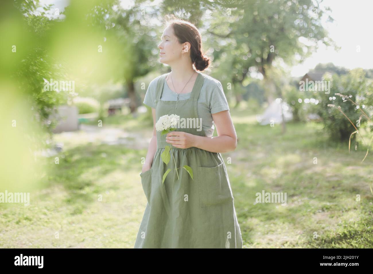 gardening and profession concept - young woman in apron holding flowers in the garden in summer ...