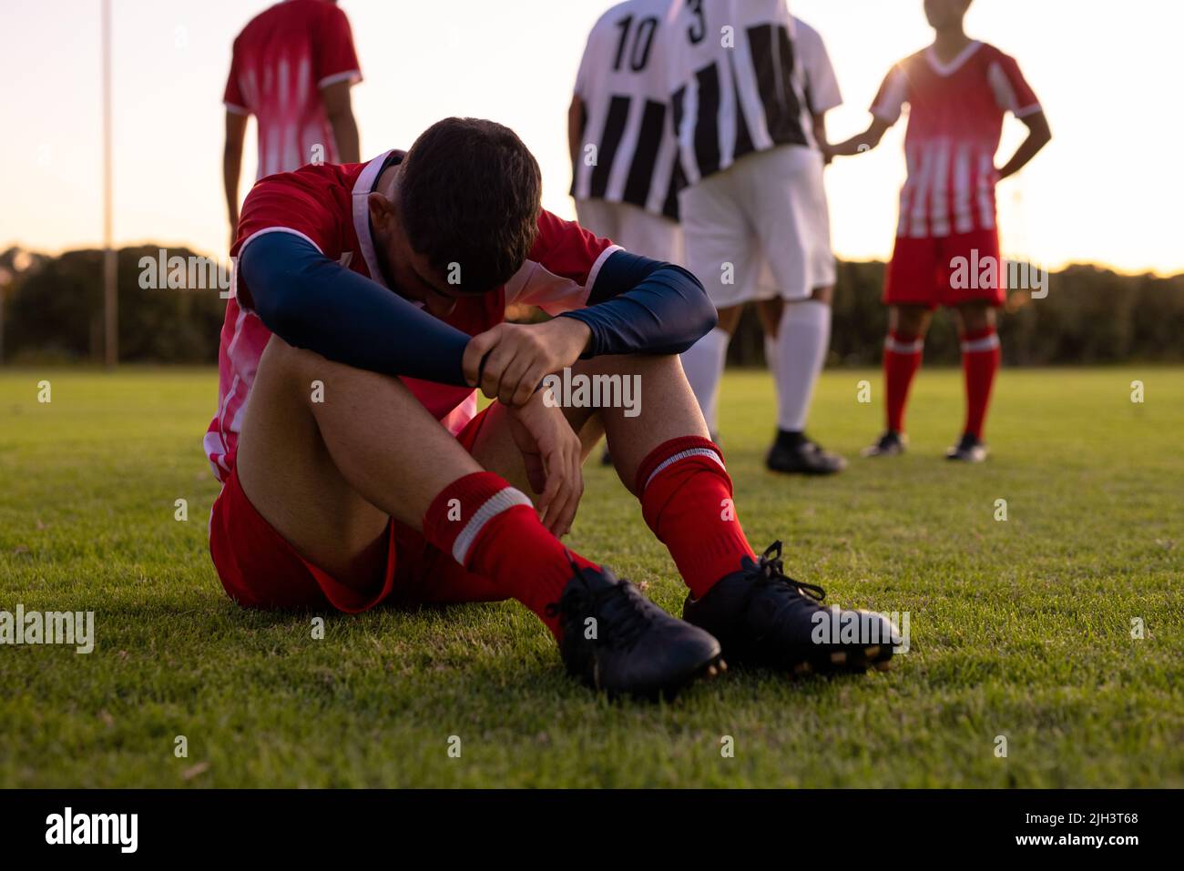 Caucasian sad male athlete sitting on grassy land with team players in ...