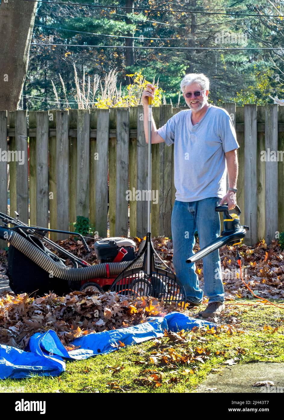 Older man plays yard warier, armed a leaf mower, rake, tarp and blower ...