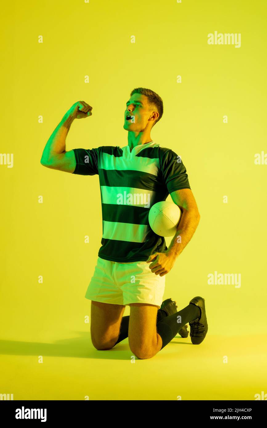 Happy caucasian male rugby player raising hands over yellow lighting ...