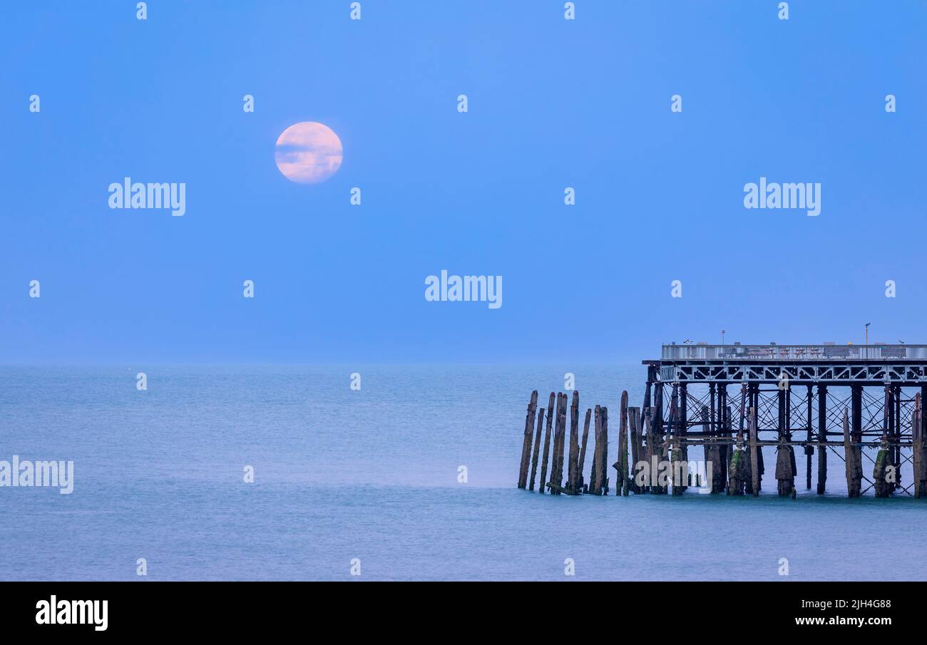 July full moon setting behind Hastings pier in East Sussex south east ...