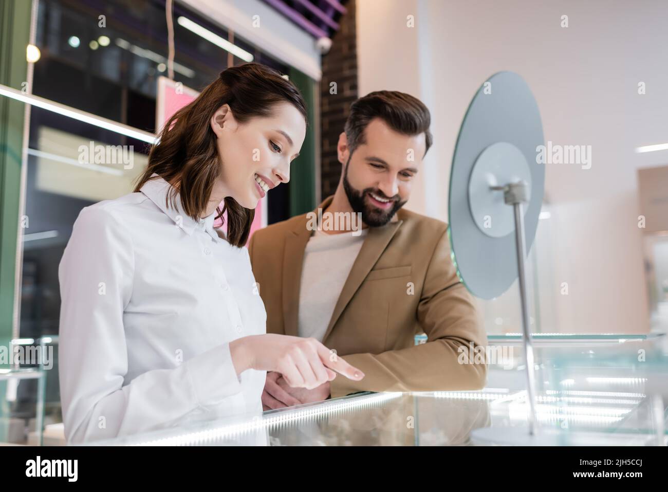 Brunette seller pointing at glass showcase near mirror and blurred ...