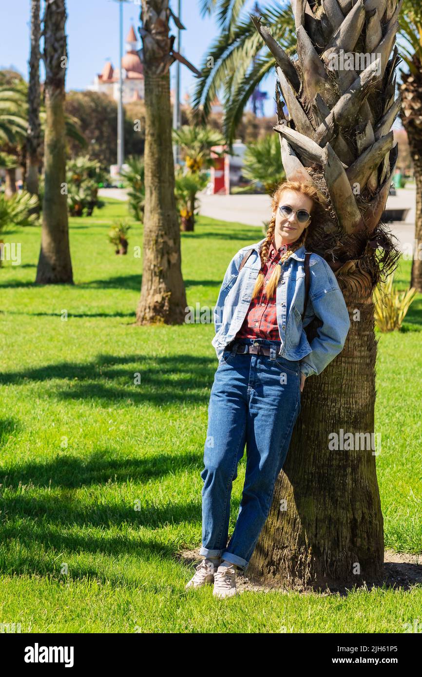 A woman in denim stands on lawn leaning on a palm tree trunk. Spring ...