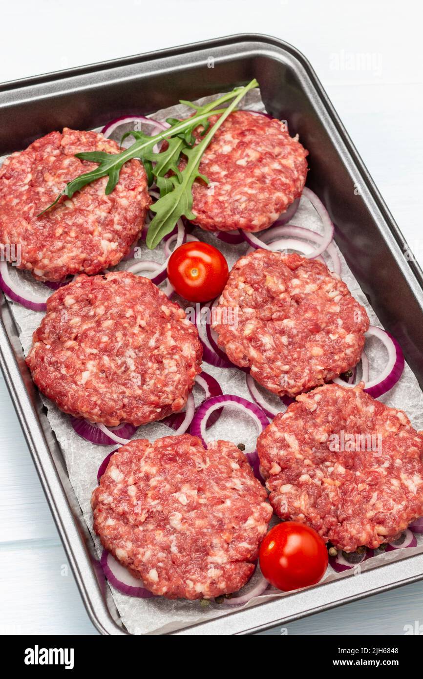Raw beef burgers with chopped onions and tomatoes on baking tray. Top ...
