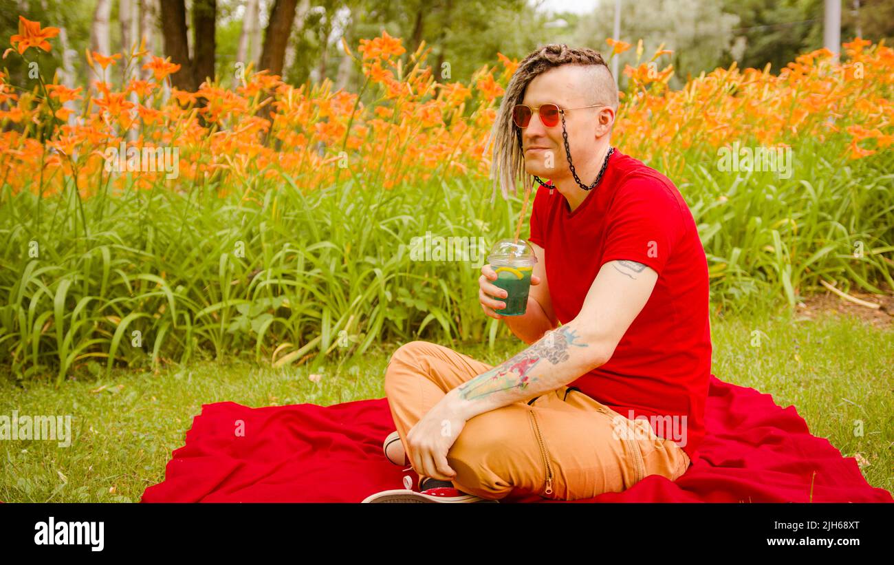 Young man drinking refreshing lemonade, sitting on rug in city park ...