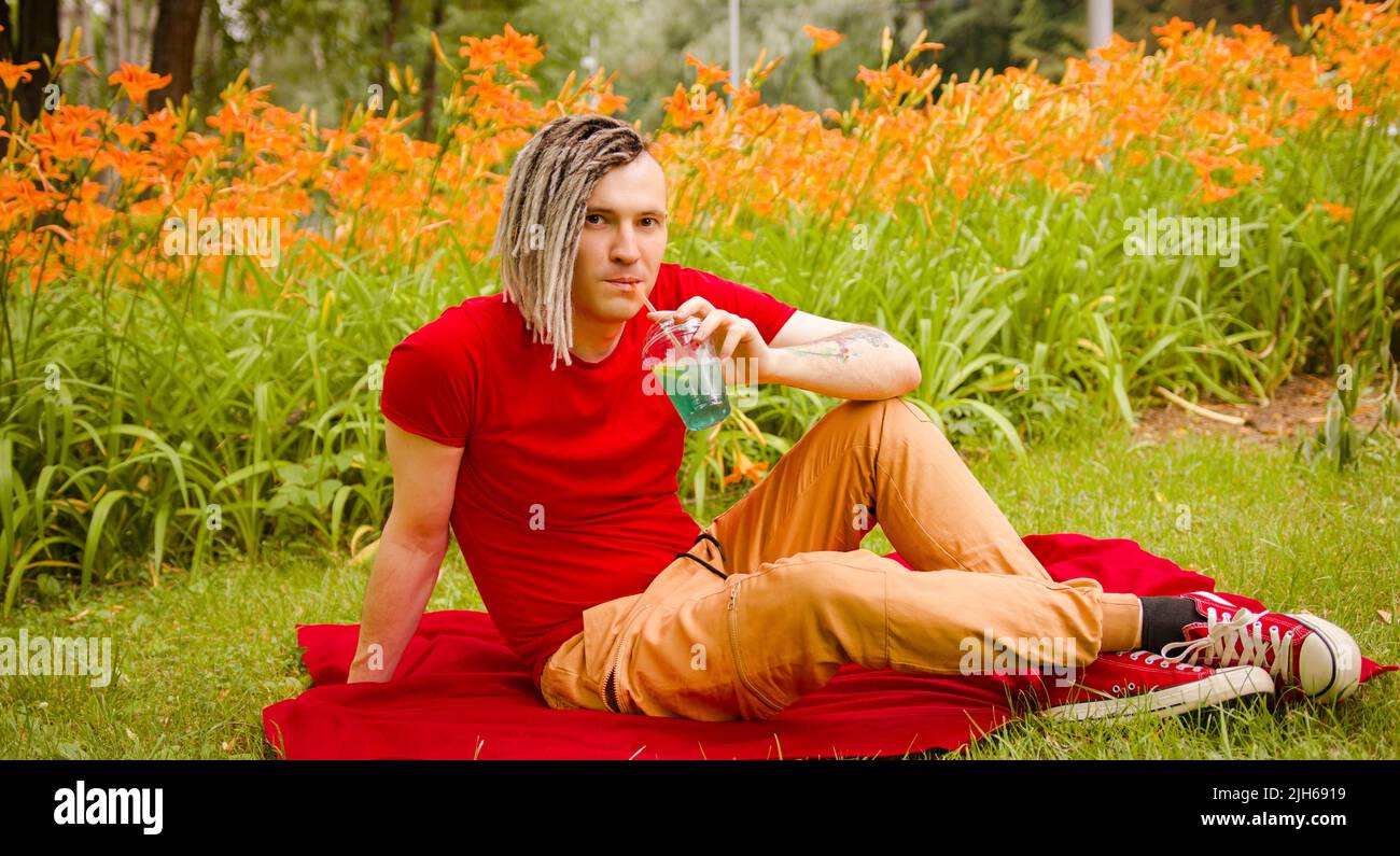 Young man drinking refreshing lemonade, sitting on rug in city park ...