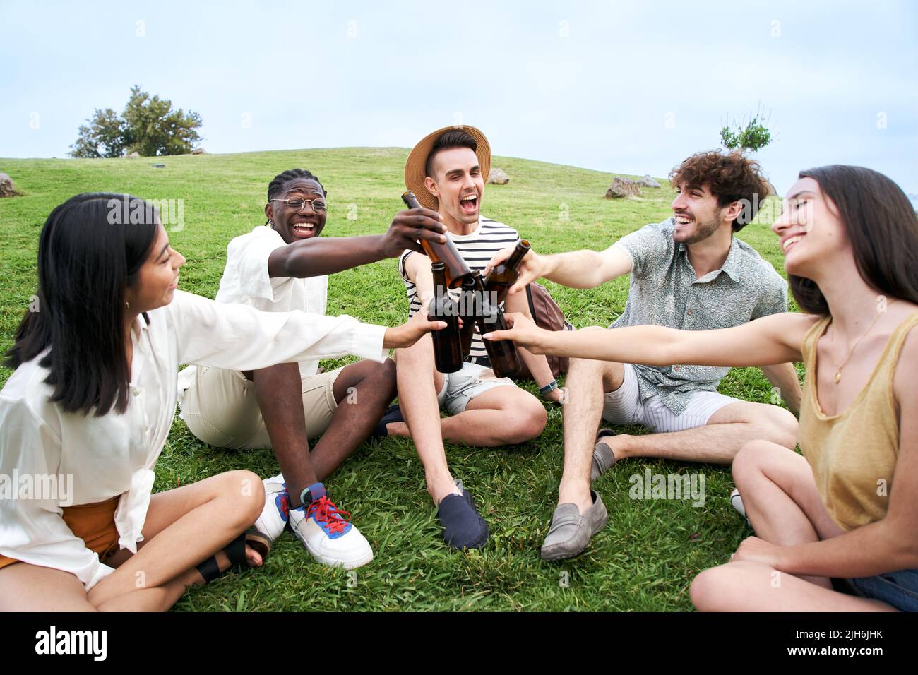 Group of young people having fun sitting at grass outside. Clinking ...