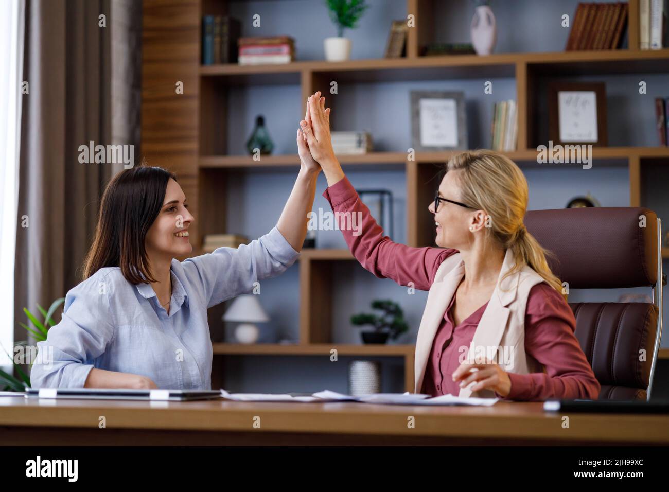 Two young smiling business women giving high-five in modern office. Happy female workers ...