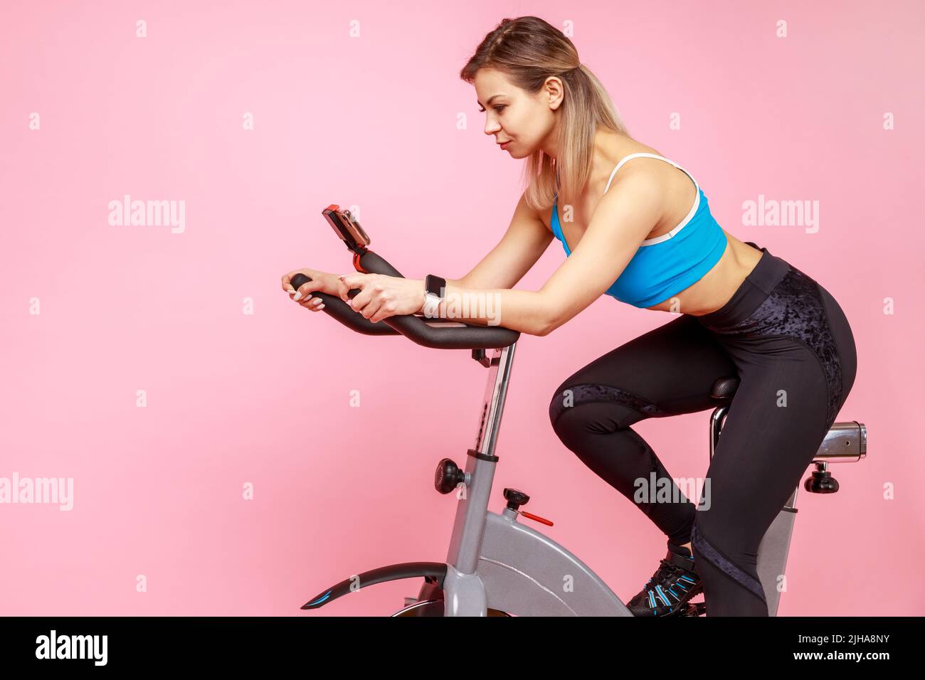 Beautiful young adult blonde woman riding exercise bike and watching ...