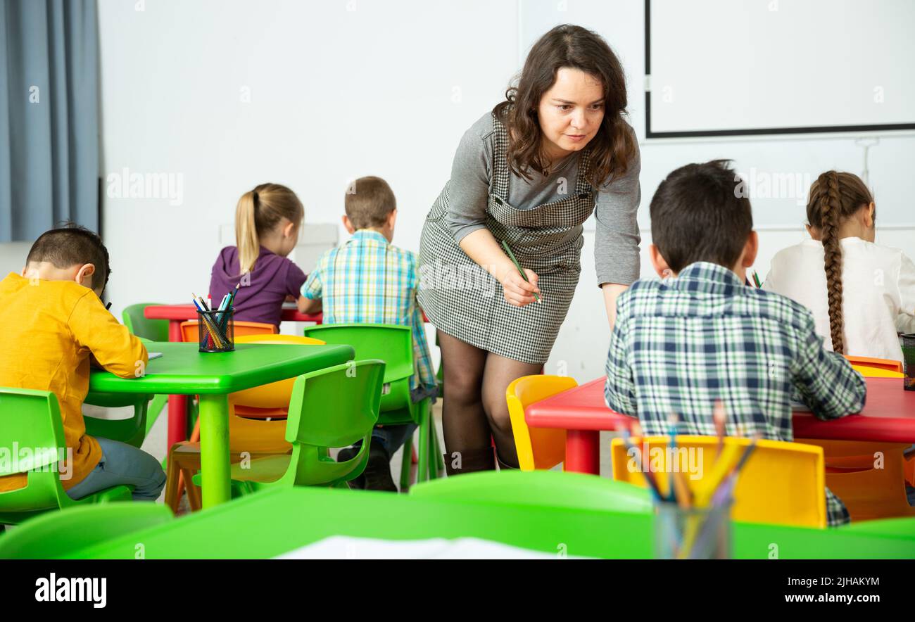 Positive boys and girls studying and female teacher helping in ...