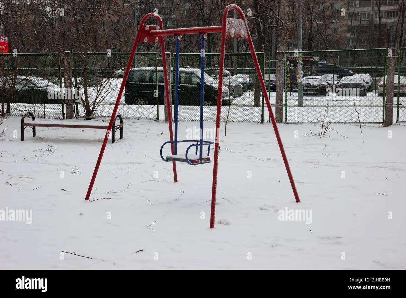 A playground in a snow-covered city: a swing, a bench, a fence, cars ...