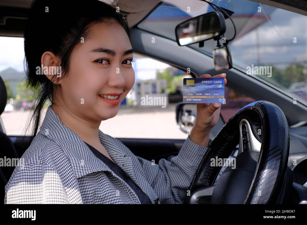 Happy beautiful Asian woman sitting inside her car showing credit card payment at a gas station ...