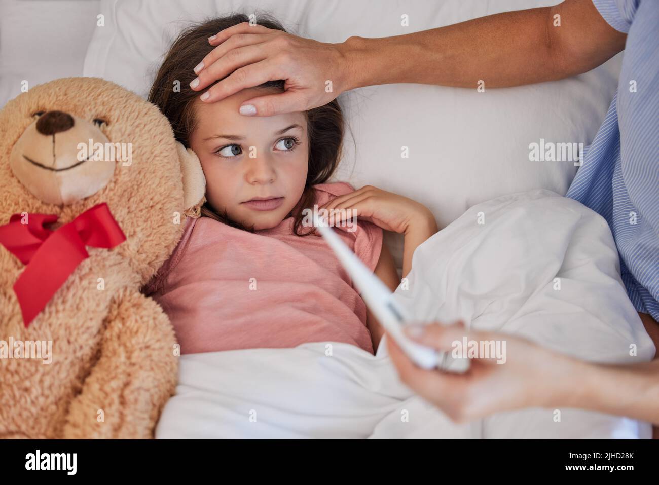 Sick little girl in bed with her teddybear while her mother uses a thermometer to check her ...
