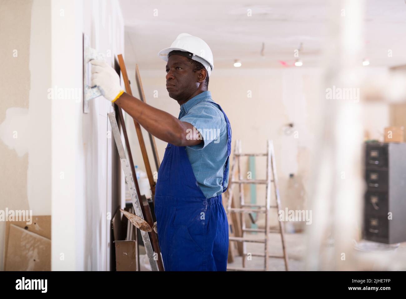 Painter in blue overalls leveling plaster on the wall with spatula ...