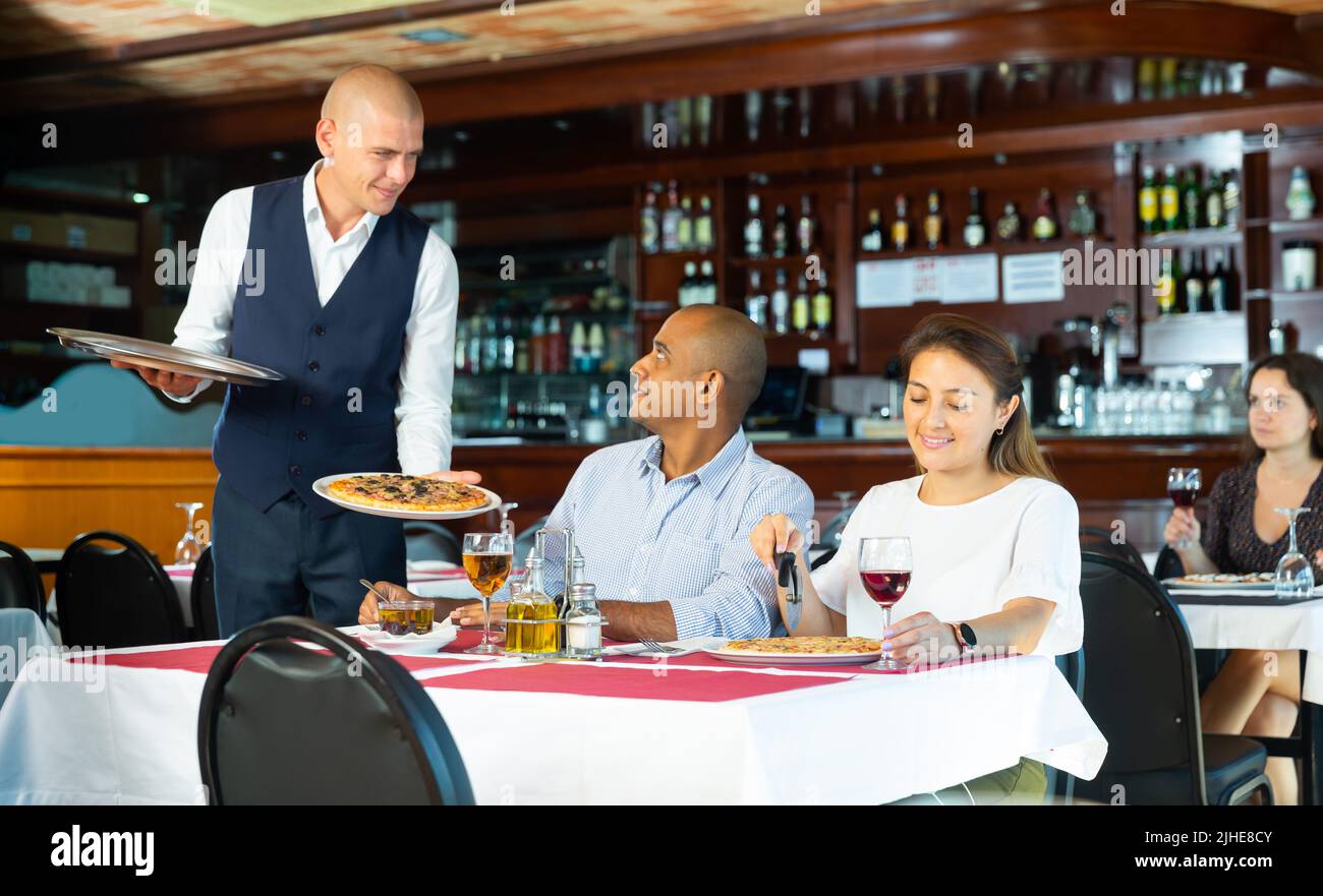 Polite waiter bringing ordered pizza to couple visited restaurant for ...