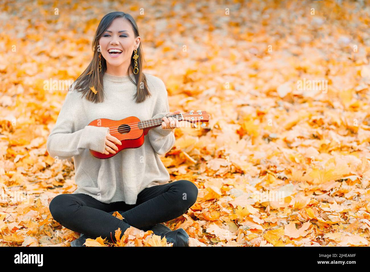 Young cheerful asian woman playing ukulele in autumn park on fallen ...