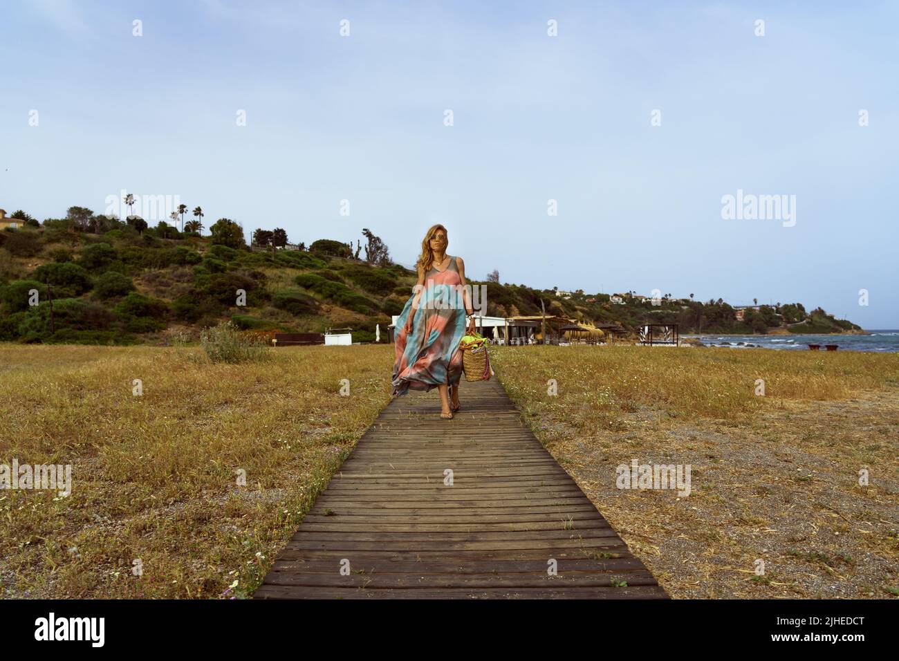 Woman in flowing dress and beach bag walking along beach promenade ...