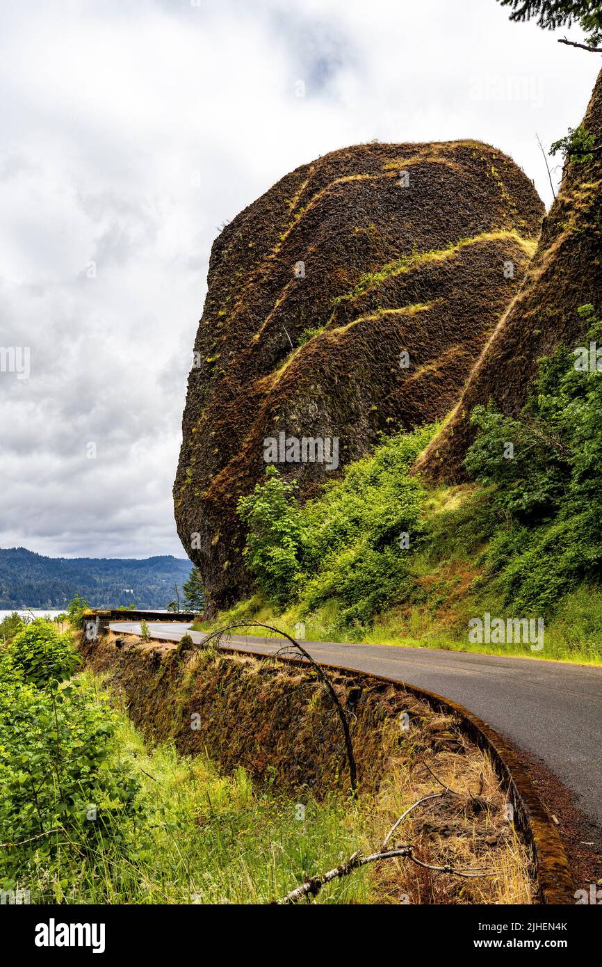 A vertical shot of a curving road beside a tall, vibrant green hill in ...