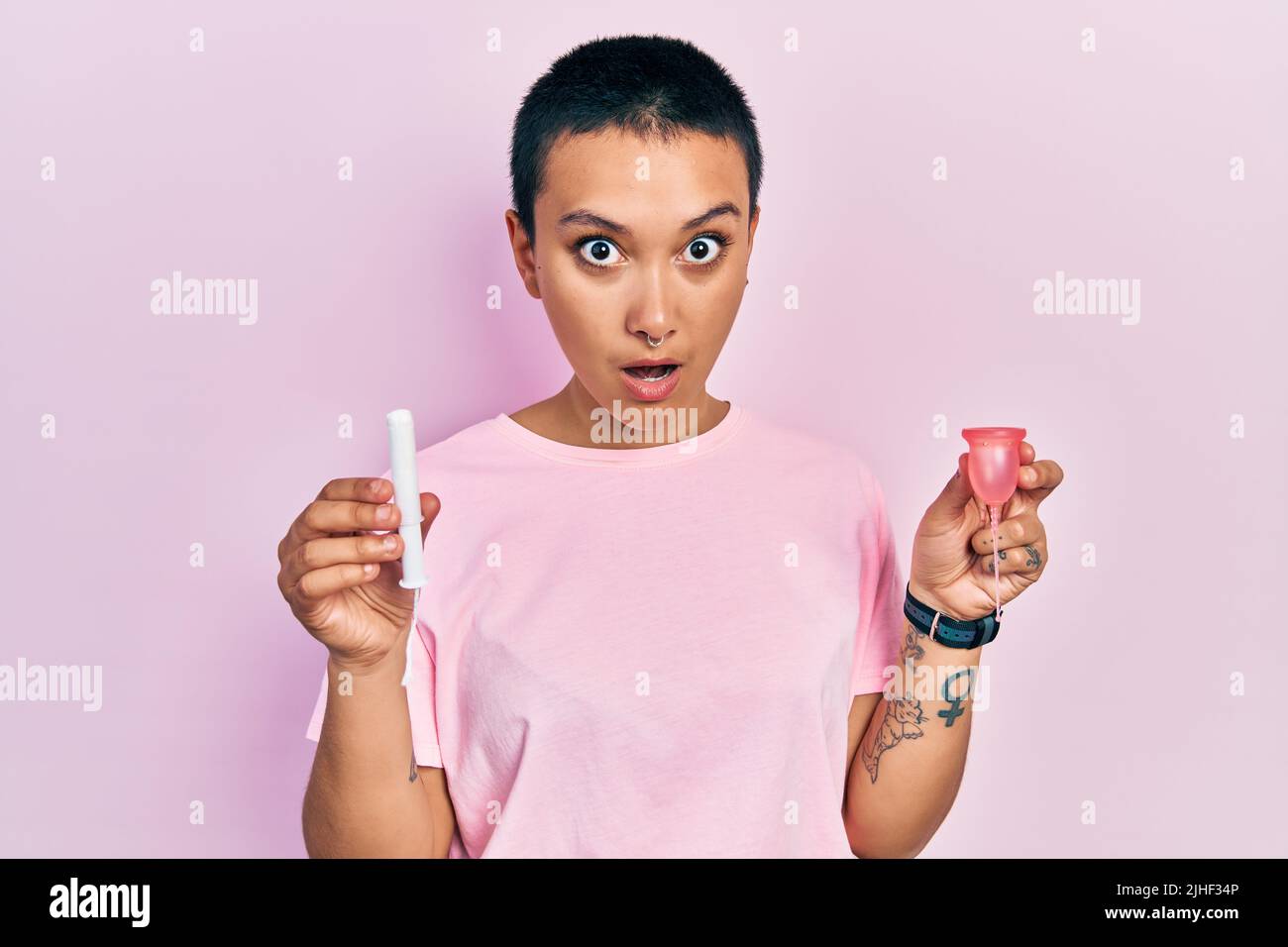 Beautiful hispanic woman with short hair holding menstrual cup and tampon in shock face, looking ...