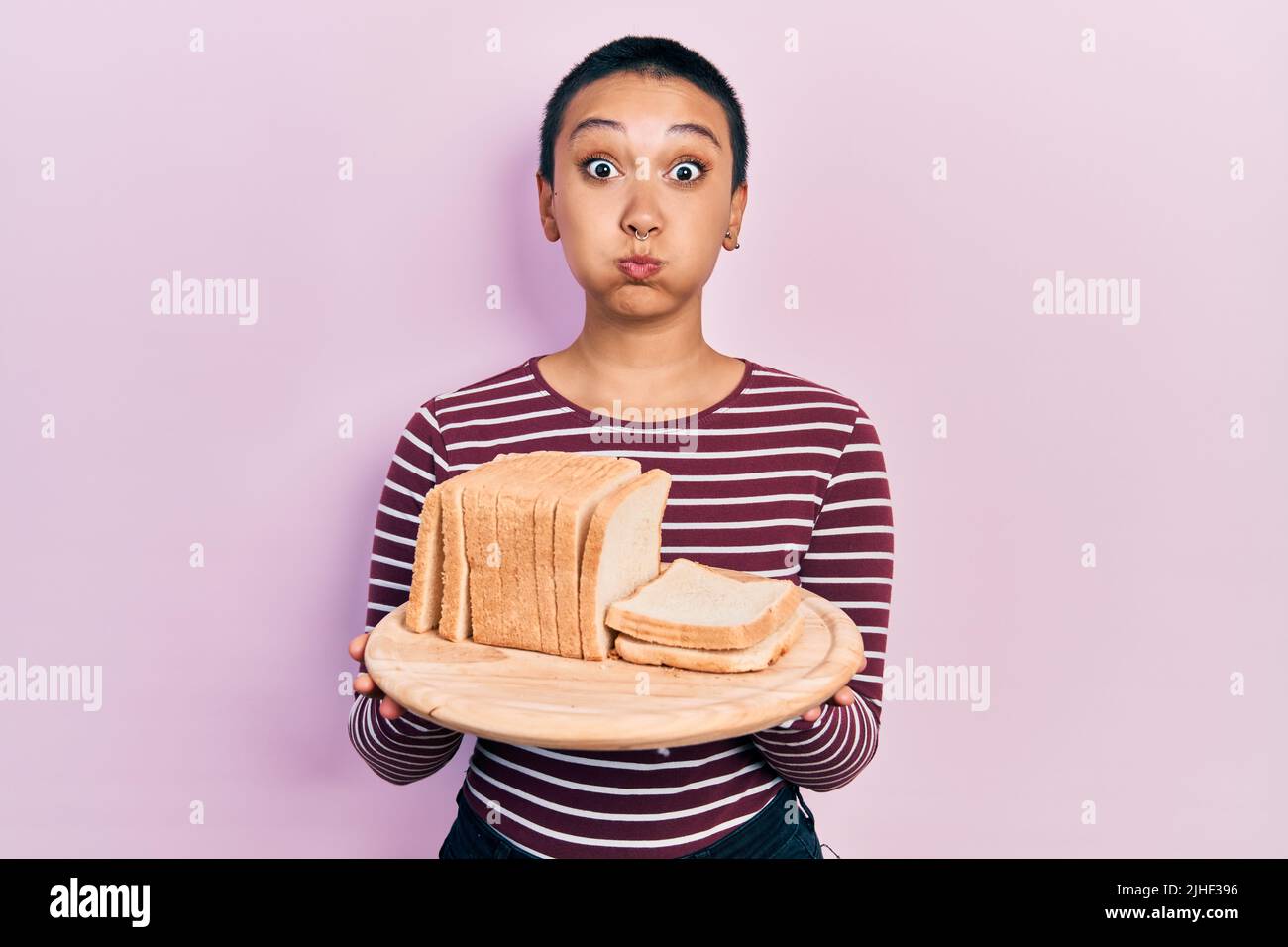 Beautiful hispanic woman with short hair holding sandwich bread puffing cheeks with funny face ...