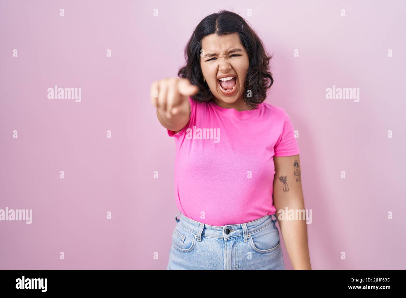 Young hispanic woman standing over pink background pointing displeased and frustrated to the ...