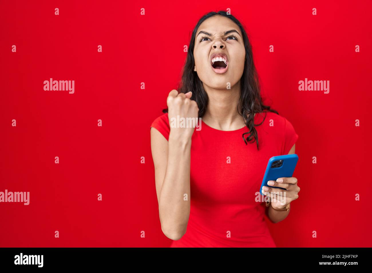 Young brazilian woman using smartphone over red background angry and ...