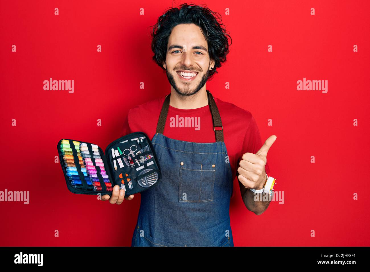 Handsome hispanic man wearing apron holding sew kit smiling happy and ...