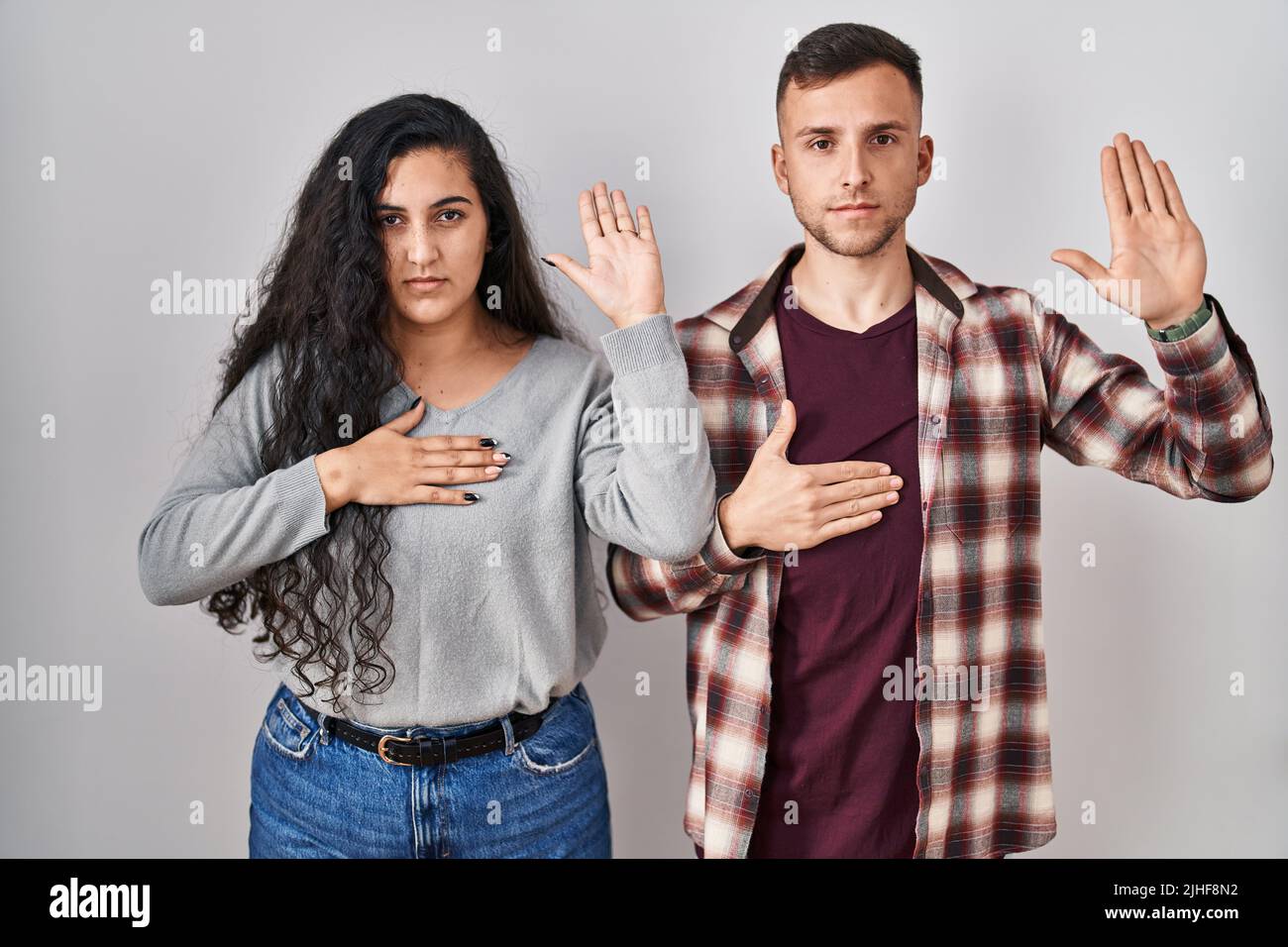 Young hispanic couple standing over white background swearing with hand ... Young hispanic couple standing over white background swearing with hand ...