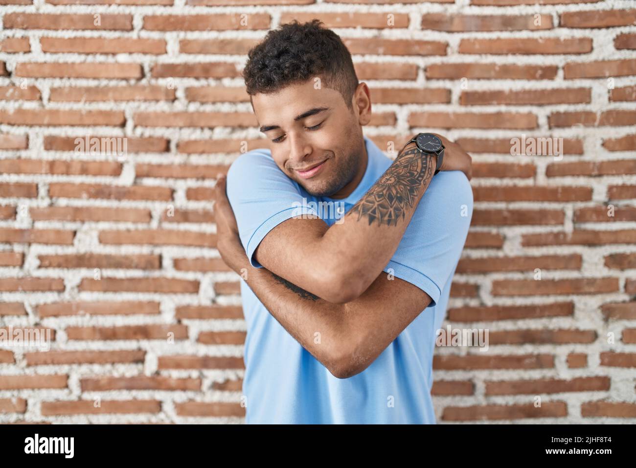 Brazilian young man standing over brick wall hugging oneself happy and ...