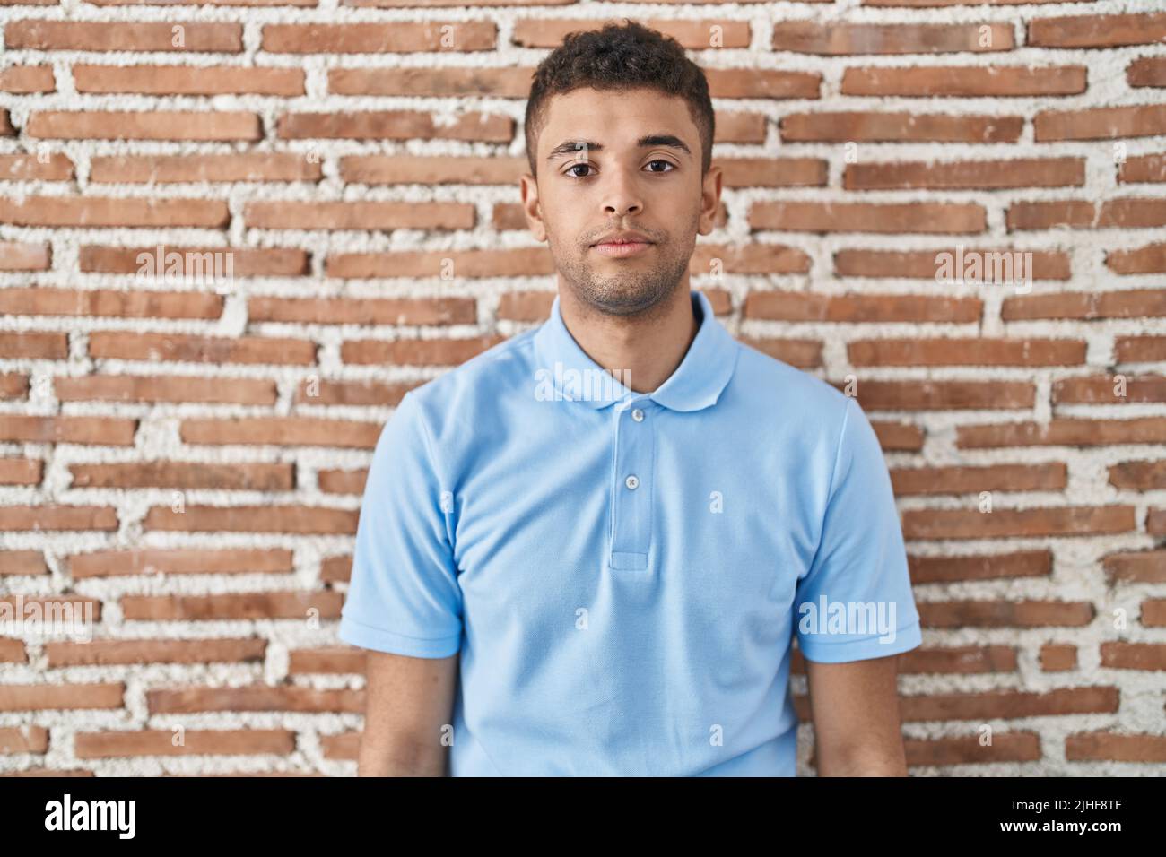 Brazilian young man standing over brick wall relaxed with serious ...