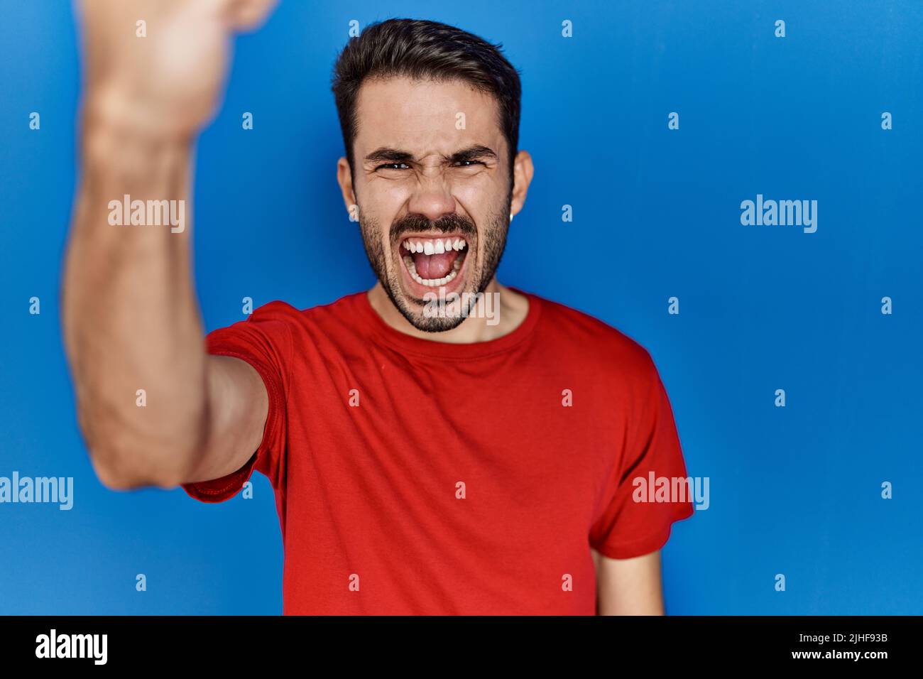 Young hispanic man with beard wearing red t shirt over blue background ...