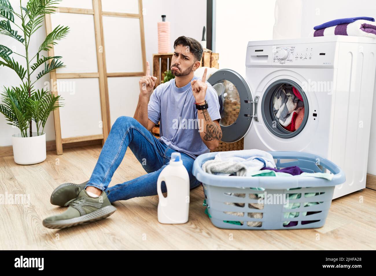 Young hispanic man putting dirty laundry into washing machine pointing ...