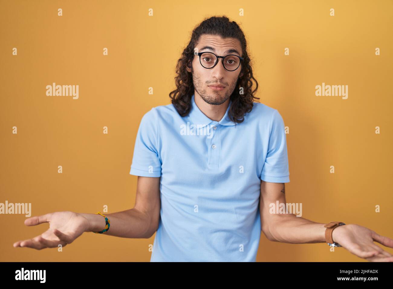 Young hispanic man standing over yellow background clueless and ...
