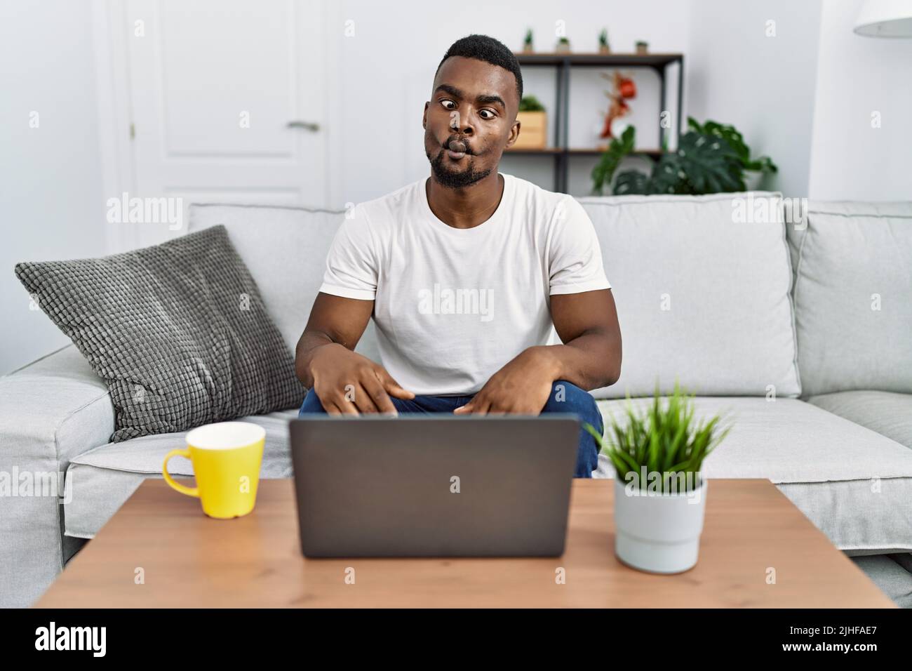 Young african man using laptop at home making fish face with lips ...