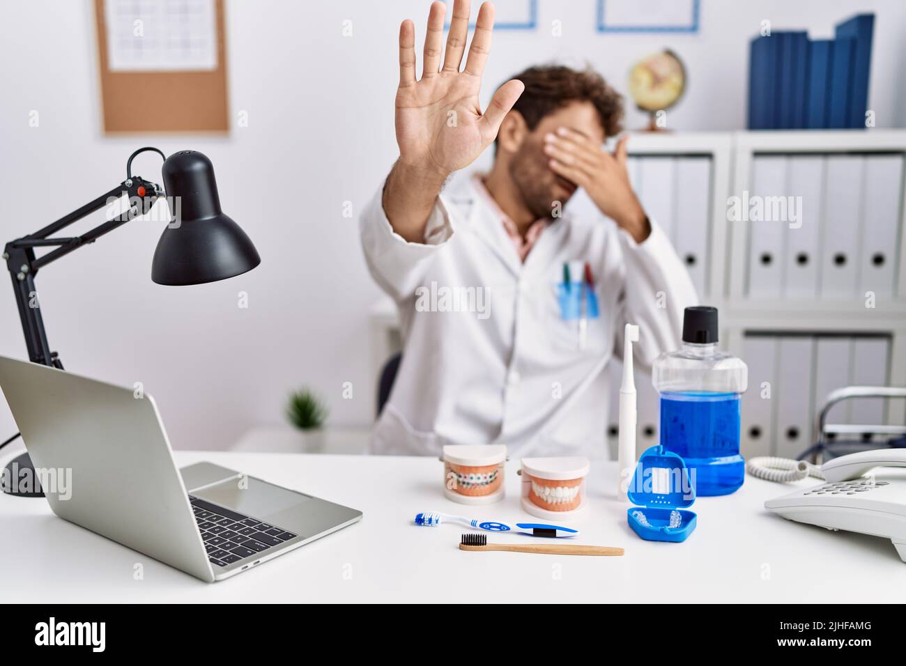 Young hispanic dentist man working at medical clinic covering eyes with ...