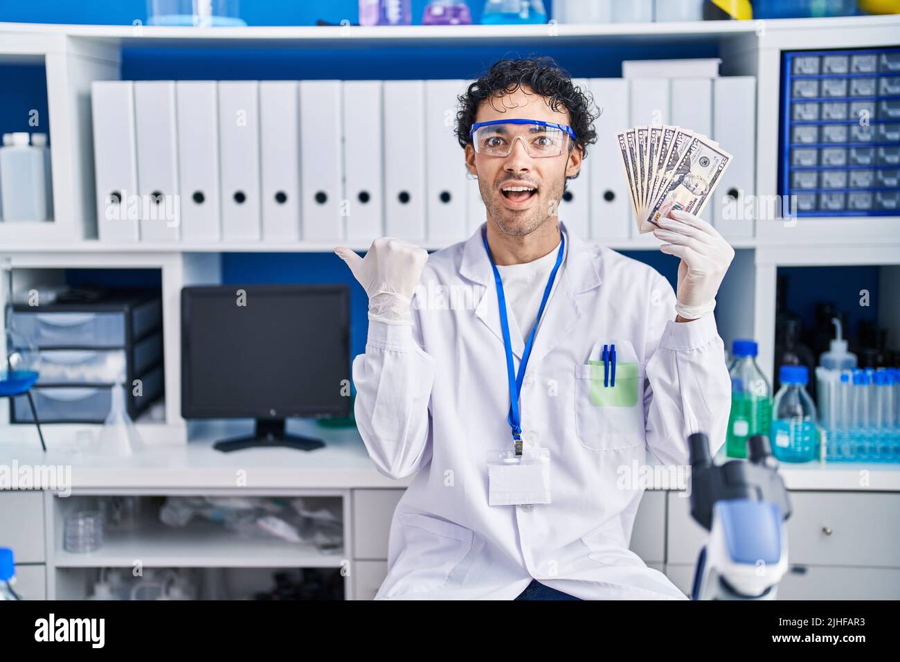 Hispanic man working at scientist laboratory holding money pointing thumb up to the side smiling ...