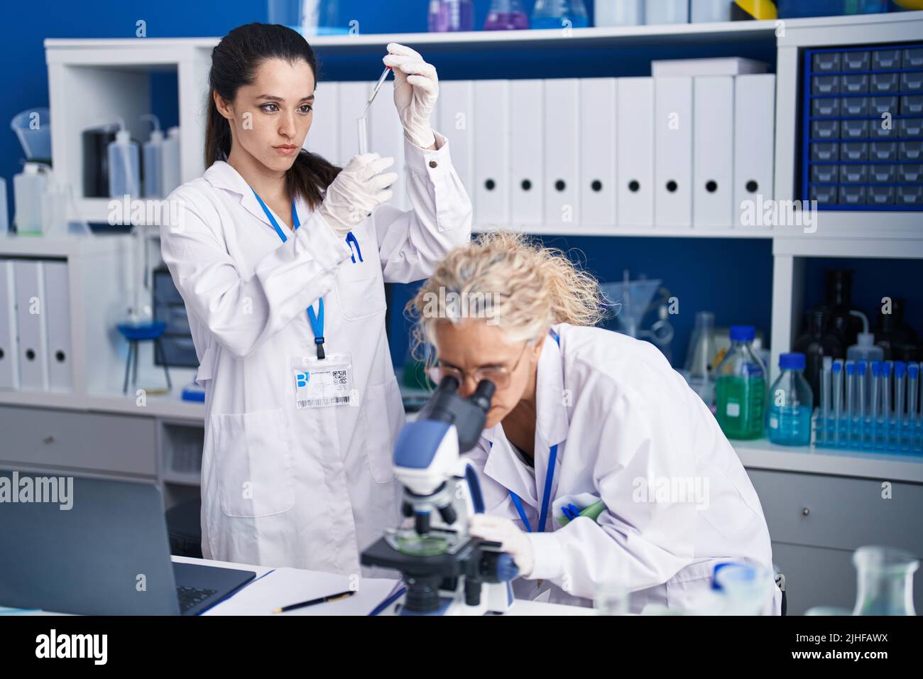 Two women scientists using microscope measuring liquid at laboratory ...