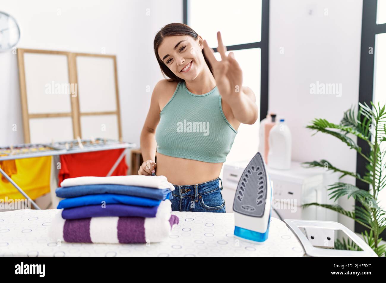 Young hispanic woman ironing clothes at laundry room smiling looking to ...