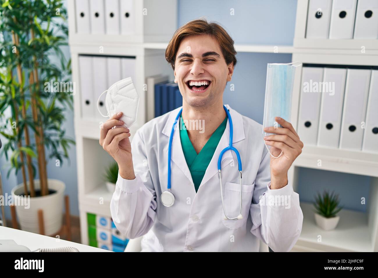 Young doctor man holding two safety mask smiling and laughing hard out ...