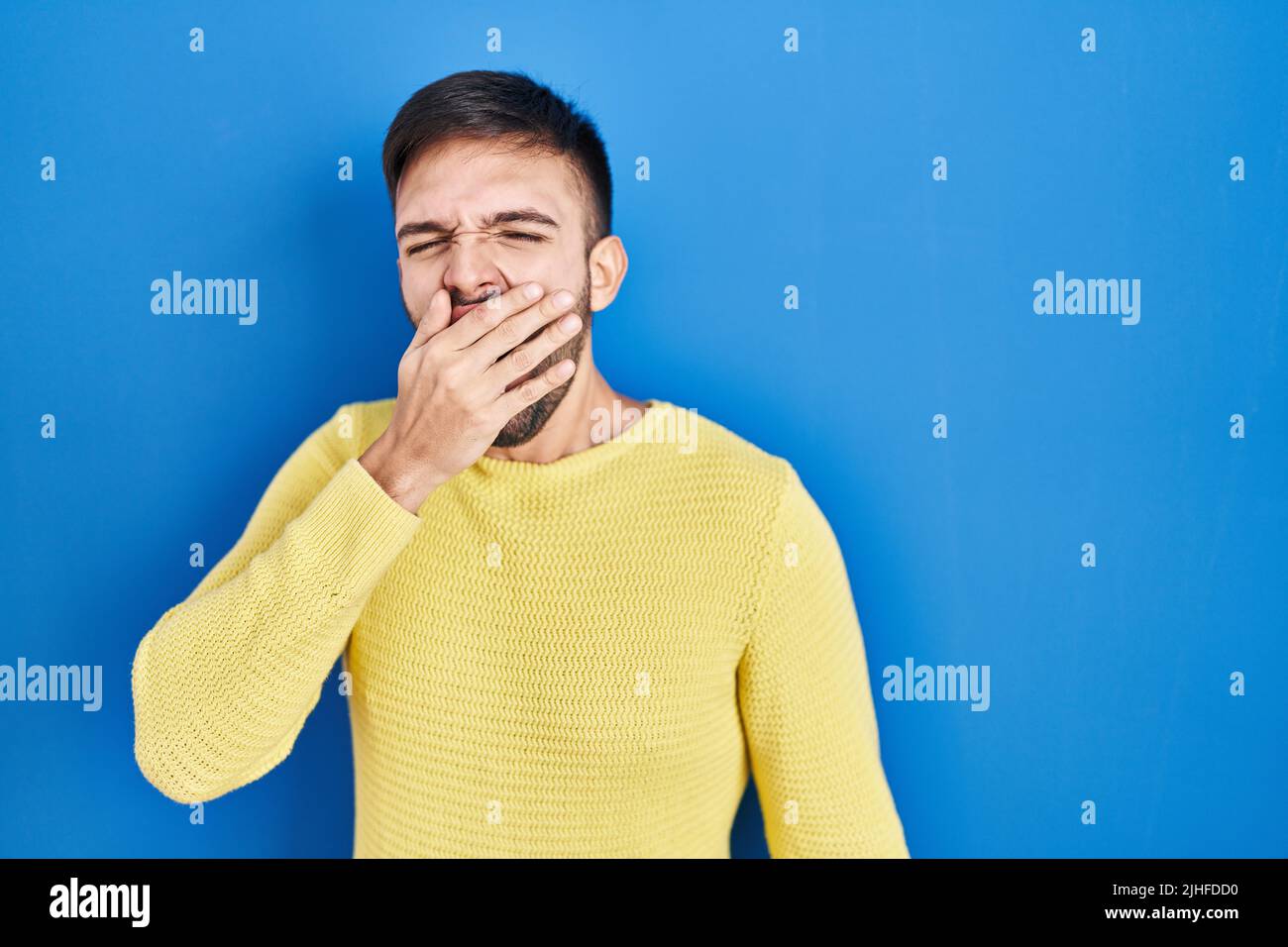 Hispanic man standing over blue background bored yawning tired covering ...
