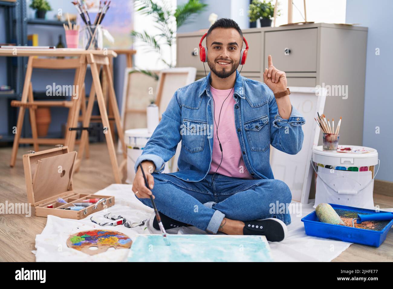 Young hispanic man painter sitting on the floor at art studio surprised ...
