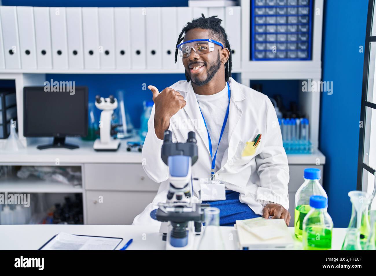 African man with dreadlocks working at scientist laboratory pointing thumb up to the side ...