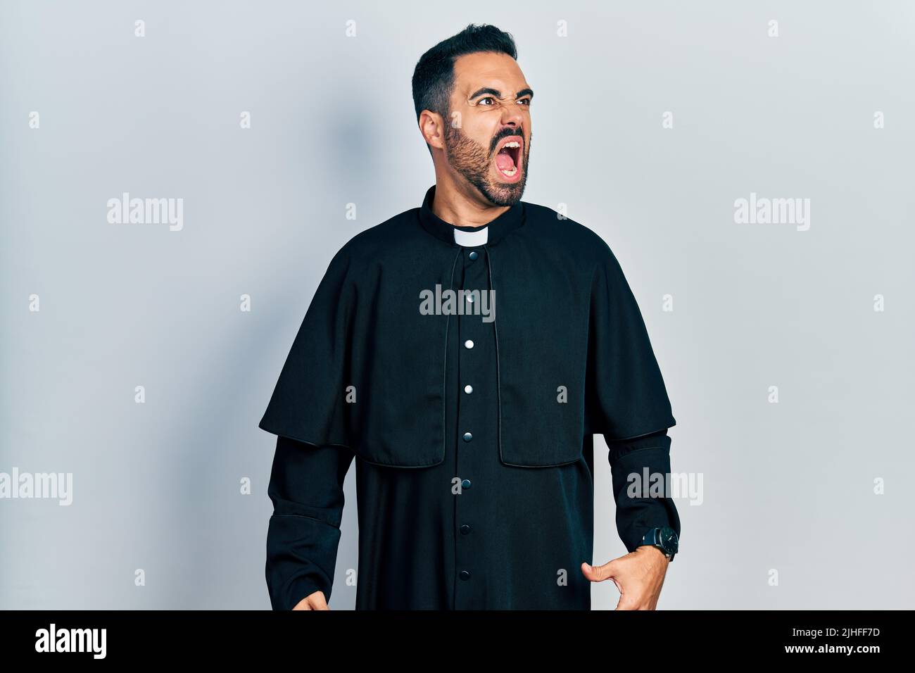 Handsome hispanic man with beard wearing catholic priest robe angry and ...