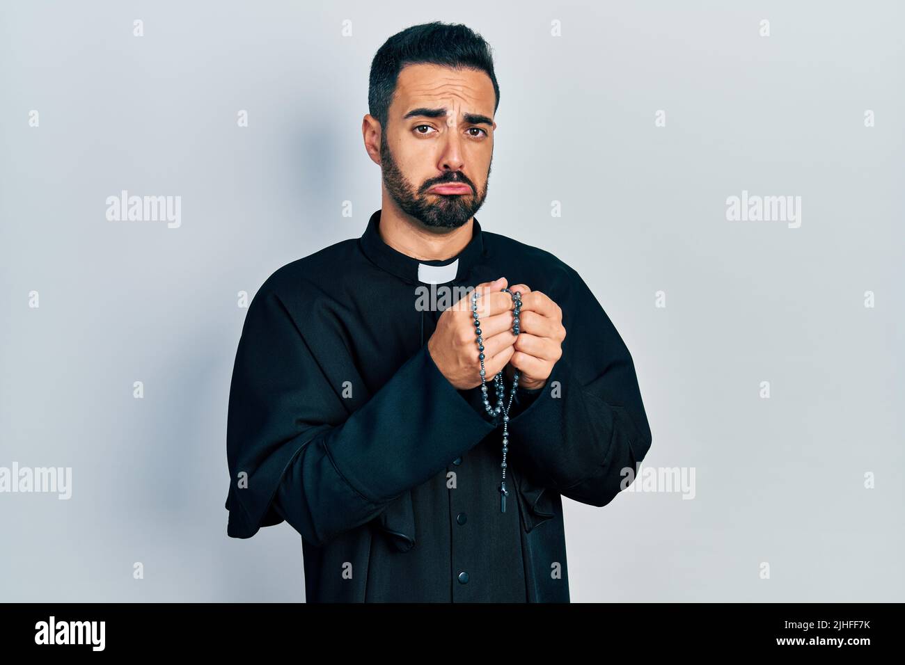 Handsome hispanic priest man with beard praying holding catholic rosary ...