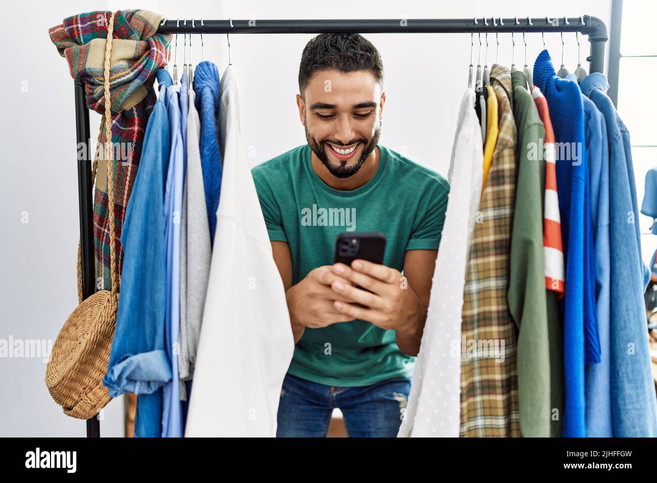 Young arab man customer using smartphone appearing through clothes rack ...