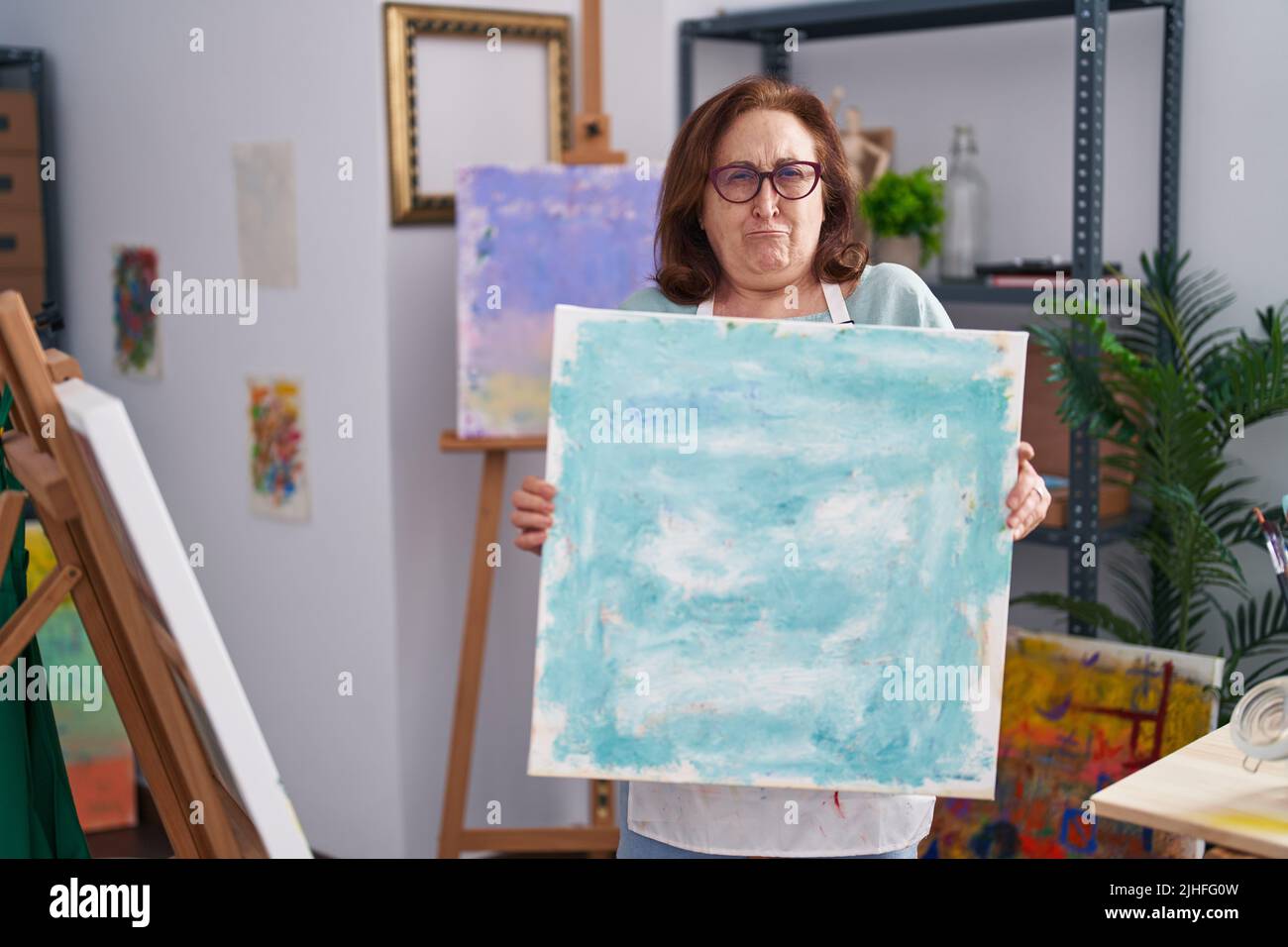 Senior woman with glasses holding canvas at art studio skeptic and ...